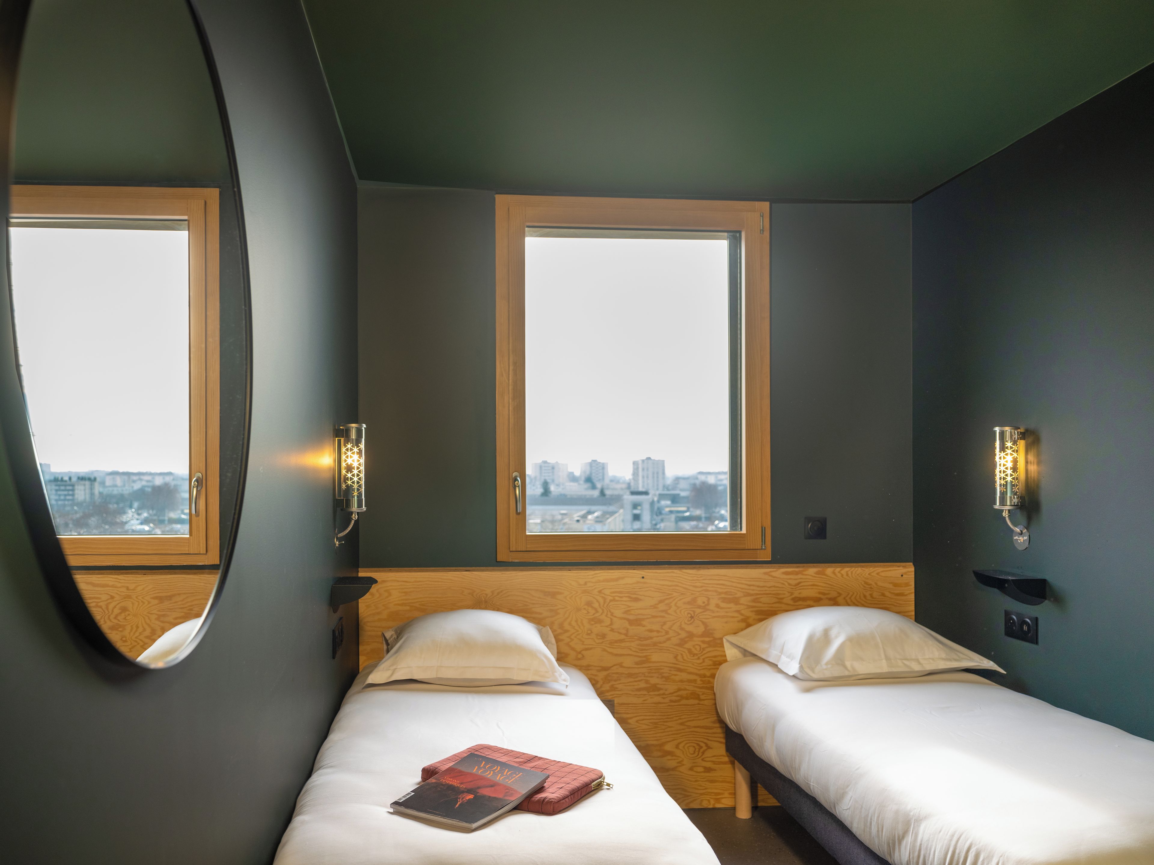 Quarto de hotel com duas camas, janelas amplas e luminárias fixadas na parede.