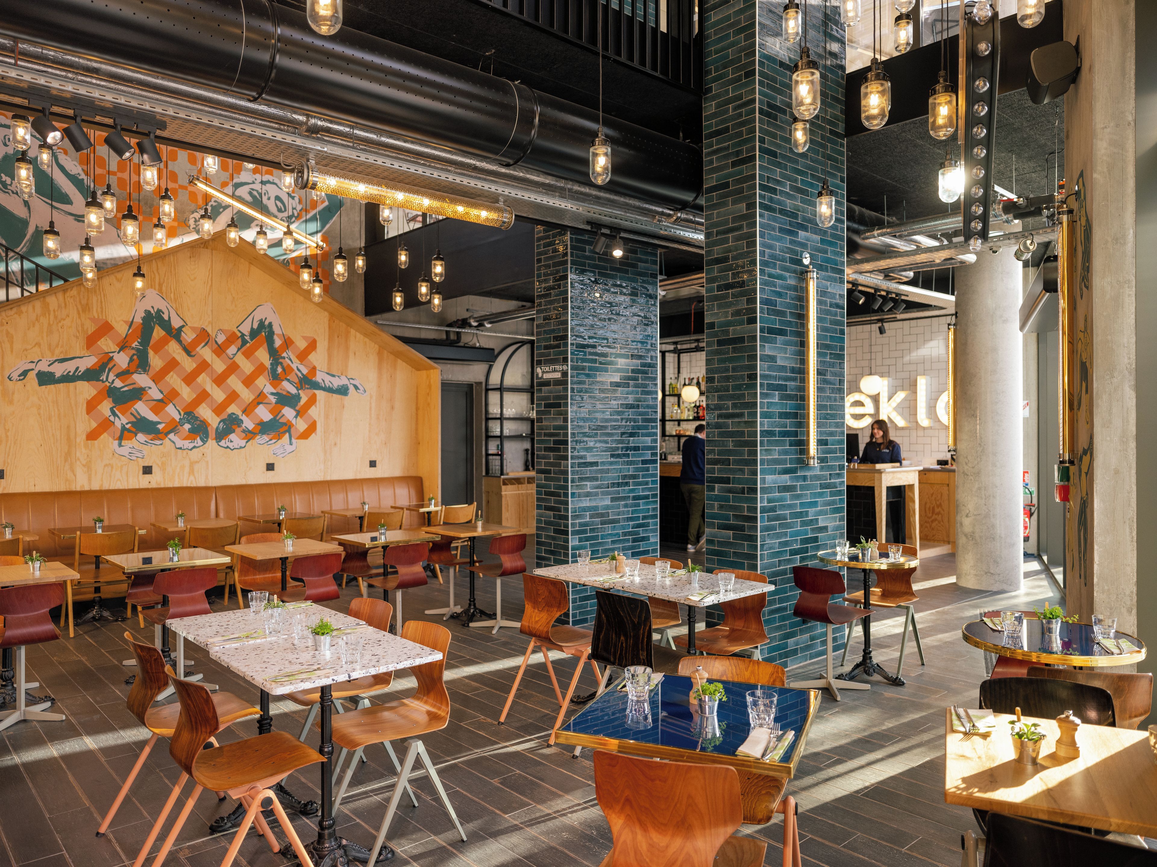 Interior de restaurante com v árias mesas e cadeiras, um bar e um mural na parede.