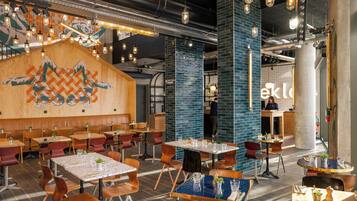 Interior de restaurante com várias mesas e cadeiras, um bar e um mural na parede.