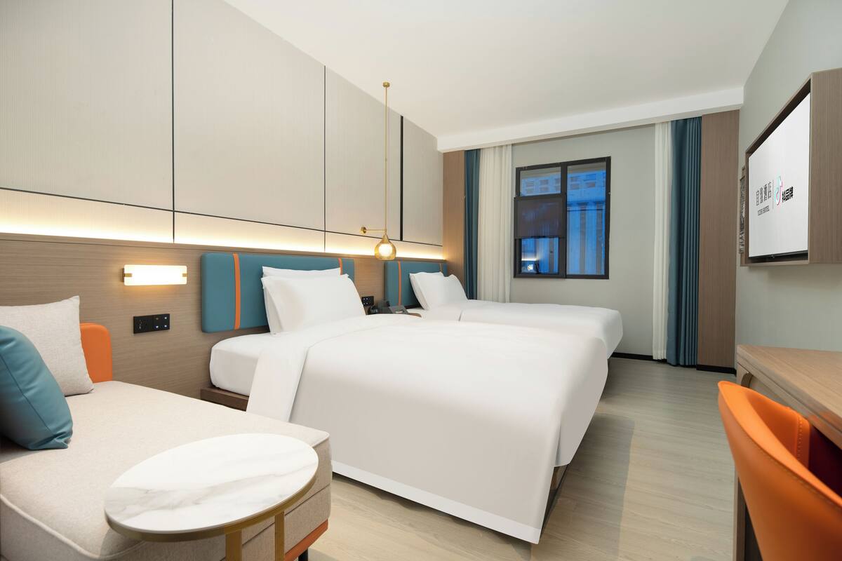 Quarto de hotel com duas camas, uma escrivaninha, uma cadeira, uma mesa redonda e uma janela grande com cortinas.