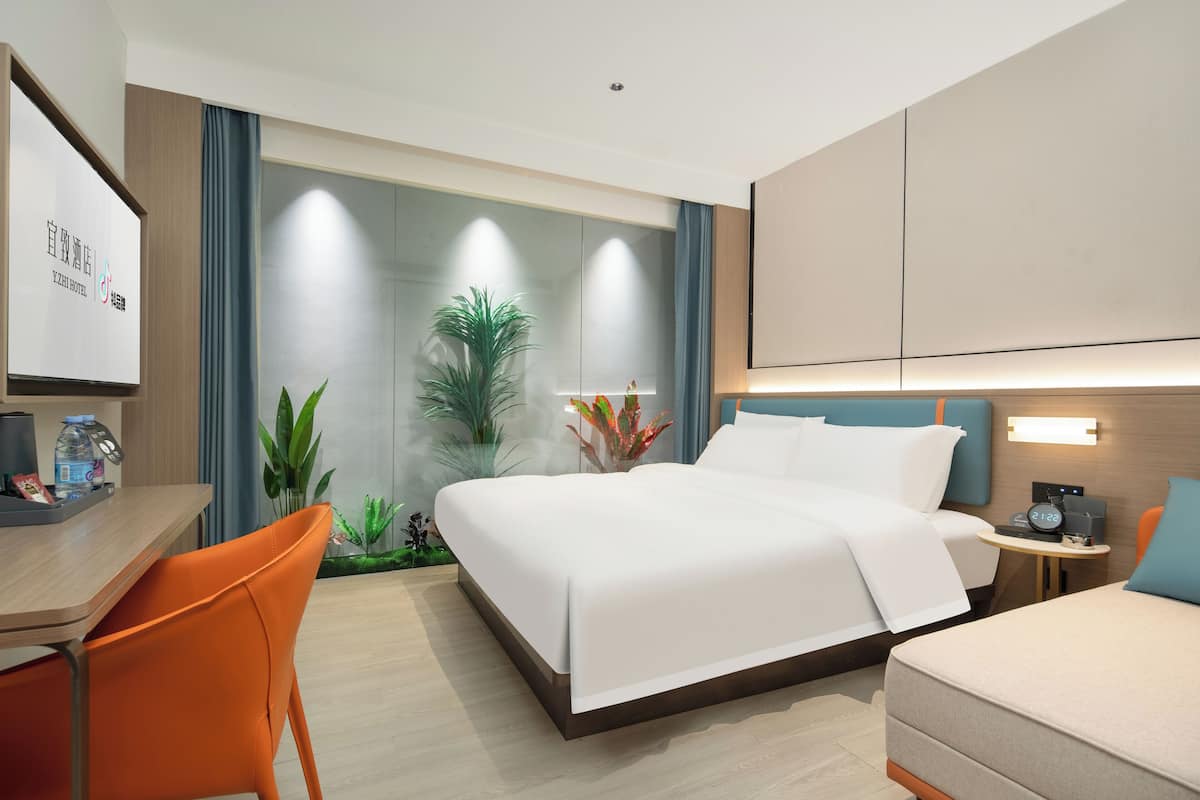 Quarto de hotel com cama, escrivaninha, cadeira, mesa de cabeceira com relógio e uma janela grande com cortinas.