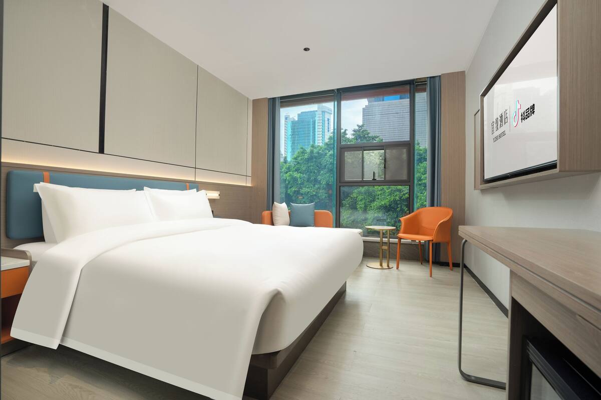 Quarto de hotel moderno com uma cama grande, uma mesa, uma cadeira e uma janela ampla com vista para a cidade.