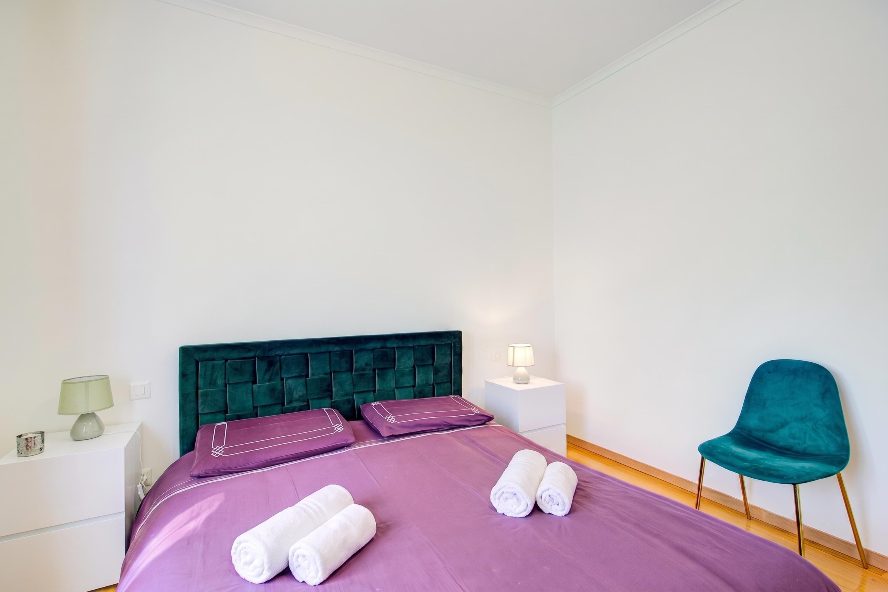 Uma cama bem arrumada com roupa de cama roxa e toalhas brancas, uma poltrona estofada em azul-esverdeado e uma mesa de cabeceira com abajur.