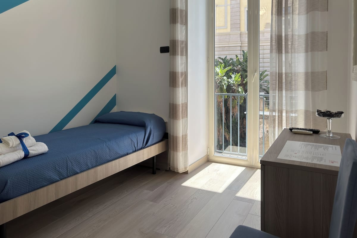 Quarto com uma cama, uma mesinha com um controle remoto e vista para uma varanda com plantas.