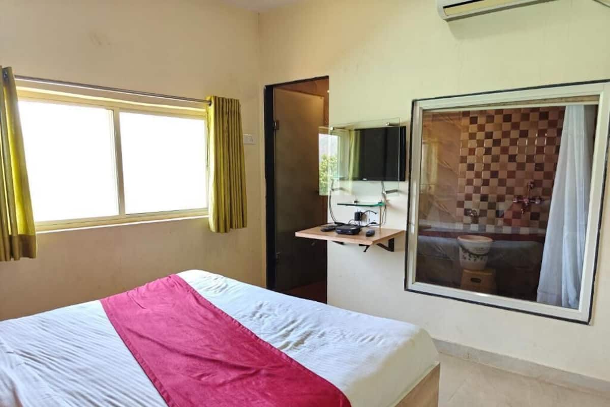 Quarto de hotel com cama, janela com cortinas, televisão montada e banheiro visível através da janela.