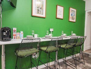 Uma área de refeições com parede verde, obras de arte emolduradas, máquina de café e banquetas de bar.
