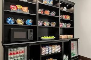 Uma cozinha compacta com micro-ondas, prateleiras repletas de lanches, um prato de maçãs verdes e uma geladeira com bebidas e produtos lácteos.