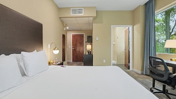 Quarto de hotel com uma cama grande, uma escrivaninha com cadeira, uma luminária, um espelho e vista para o exterior.