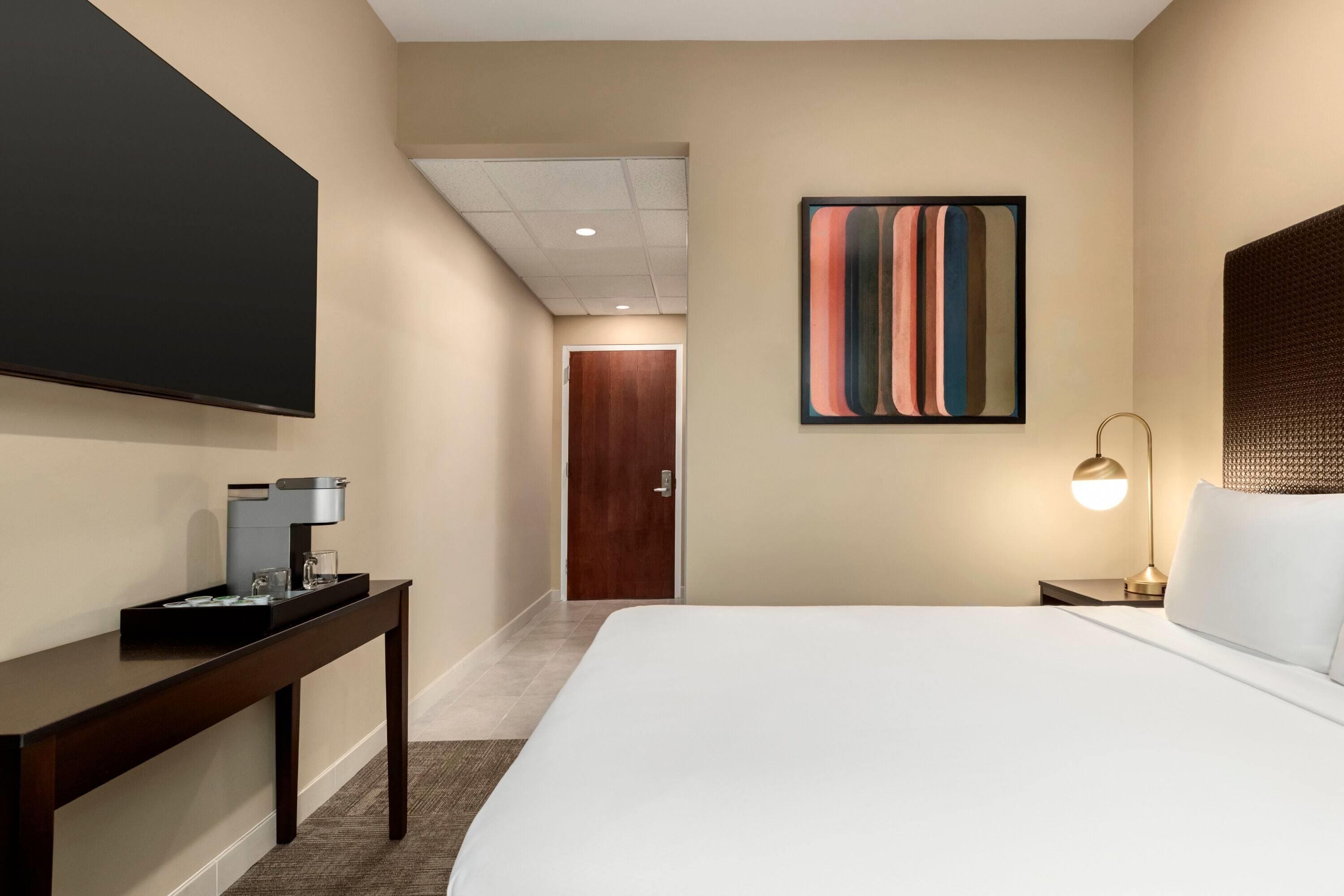 Quarto de hotel com cama, escrivaninha com cafeteira, televisão, quadro e abajur.
