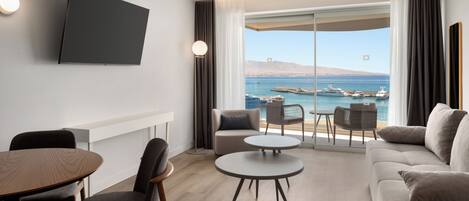 Quarto de hotel moderno com televisão de tela plana, área de refeições, sofá e vista para o mar e barcos.