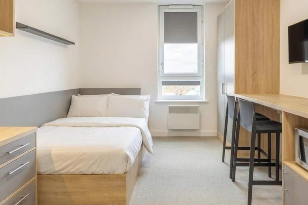 Quarto compacto com cama, escrivaninha, cadeira, televisão e janela.