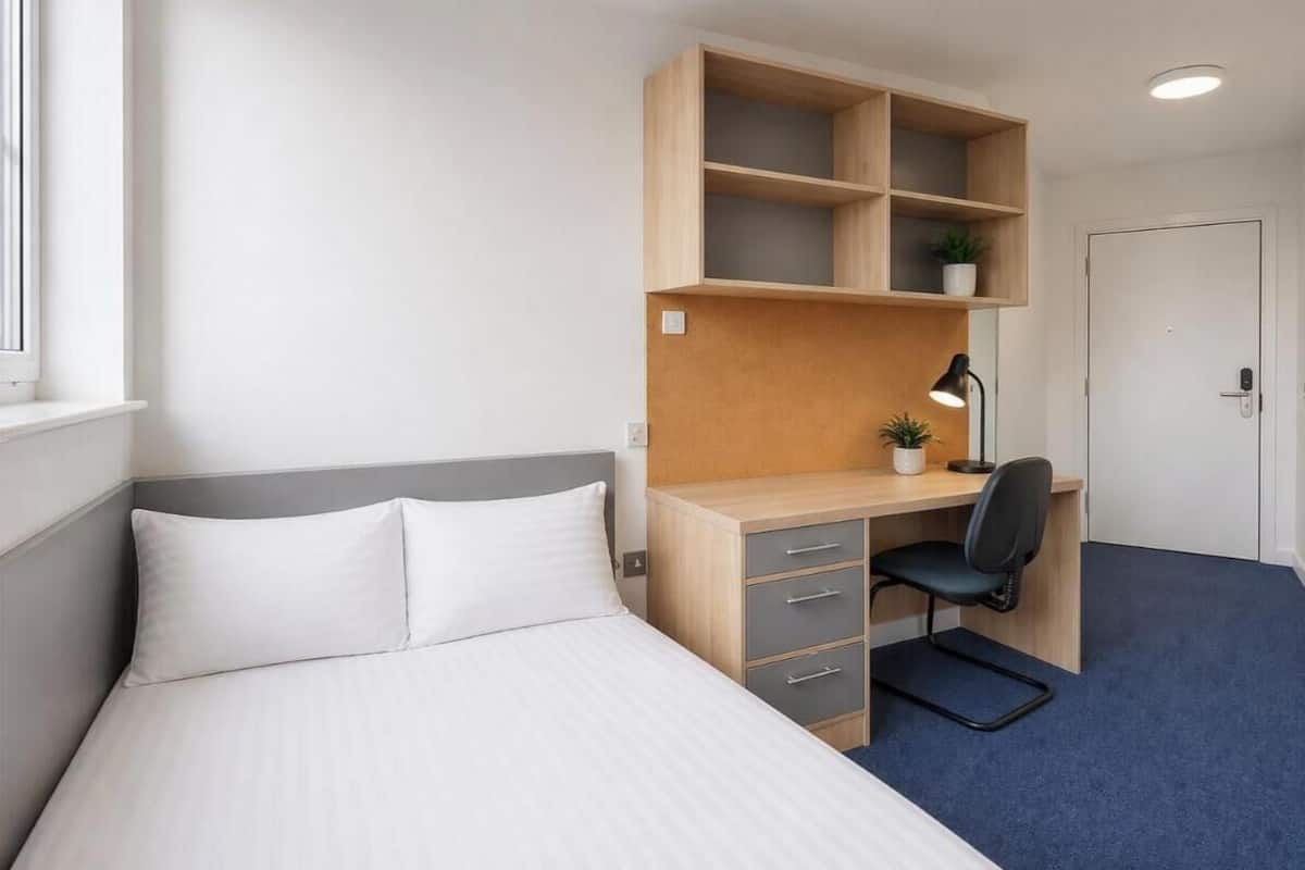 Quarto de hotel com cama, escrivaninha com gavetas, cadeira, luminária e porta.