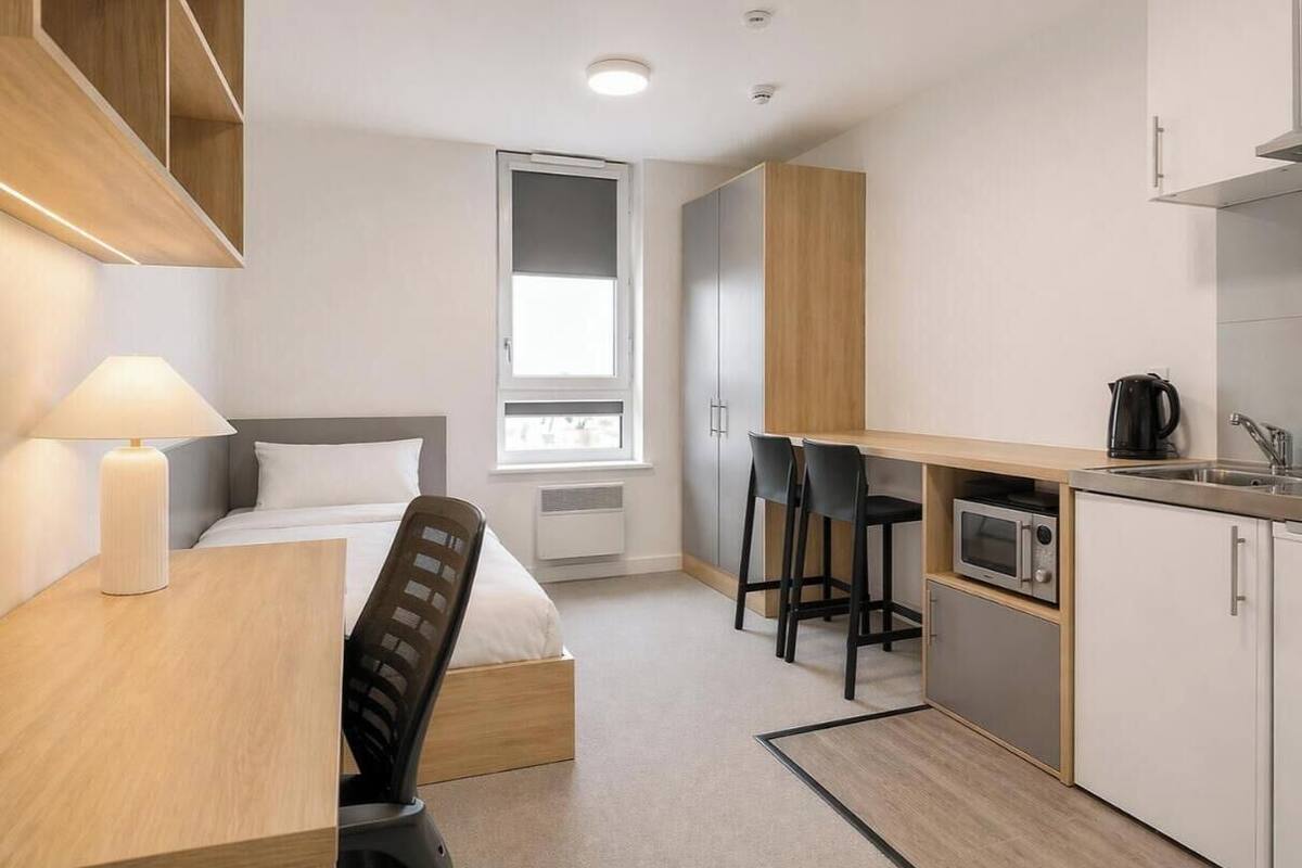 Quarto de hotel compacto com cama, escrivaninha, cozinha compacta e janela.
