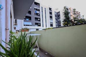 Uma varanda com um vaso de planta, uma mesa branca e vista para um edifício moderno com várias varandas.