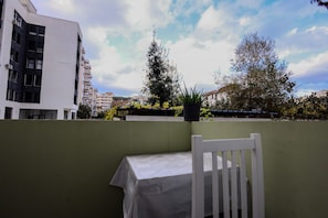 Varanda com mesa e cadeira brancas, com vista para um horizonte urbano com edifícios e árvores.