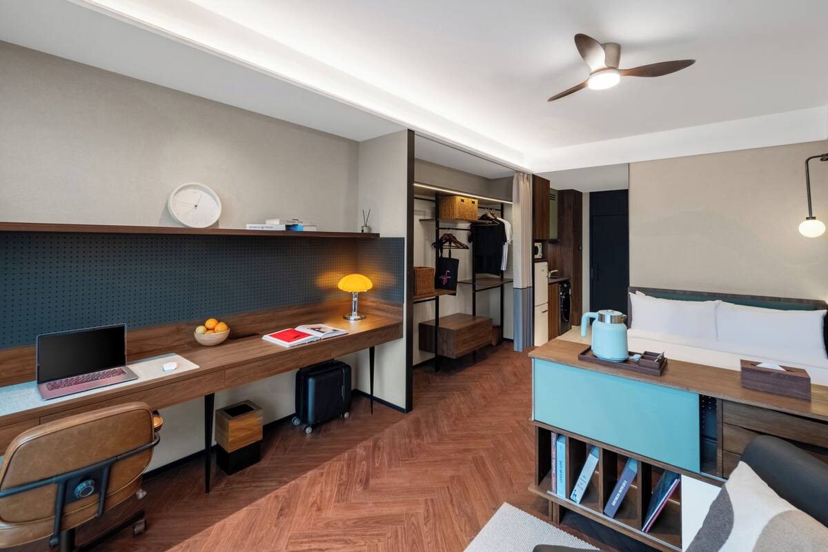 Quarto de hotel com escrivaninha, laptop, cadeira, cama, mesa de cabeceira, abajur e armário.