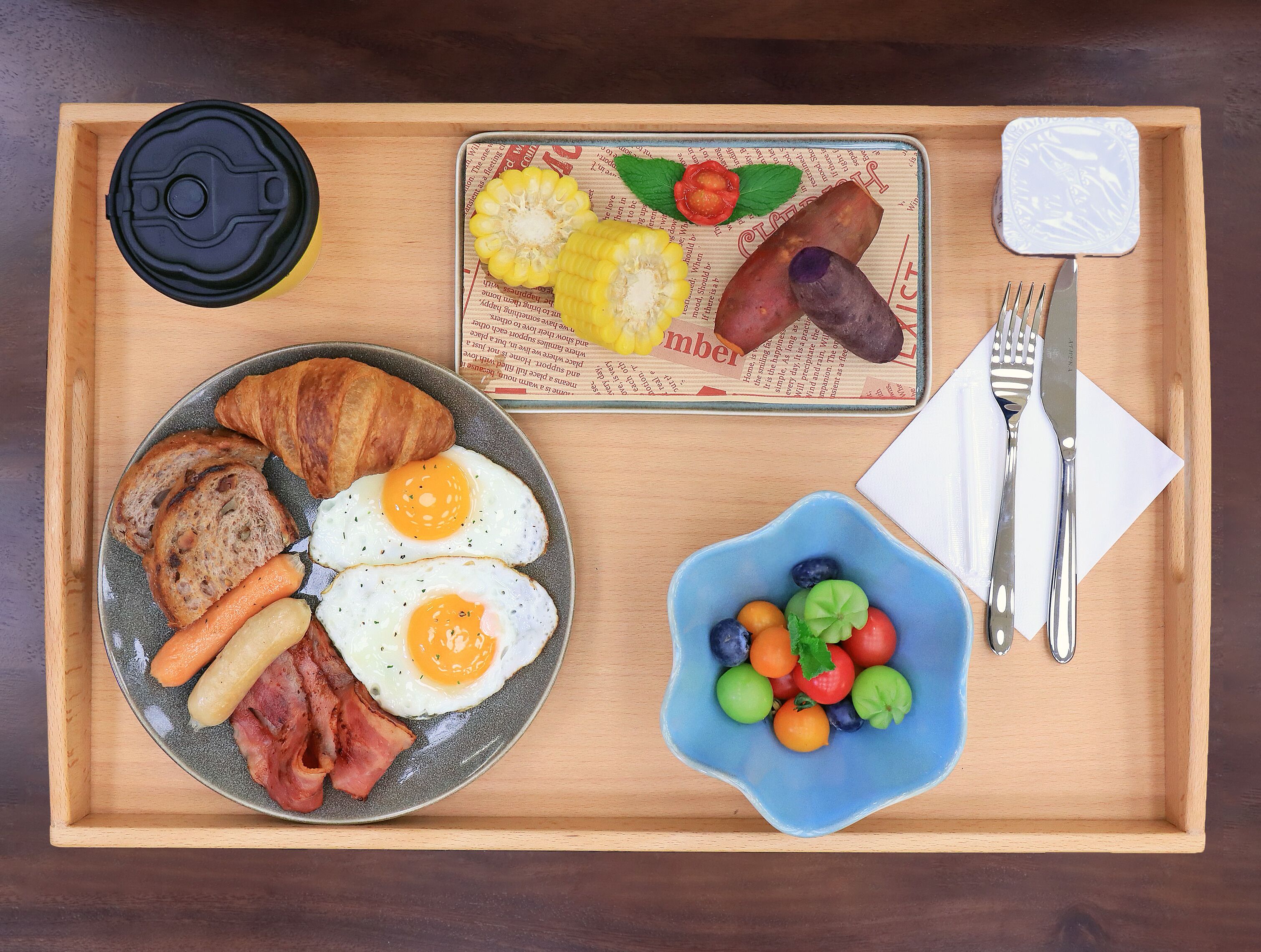 Bandeja de café da manhã com ovos, bacon, torradas, linguiça, milho e uma tigela de frutas.