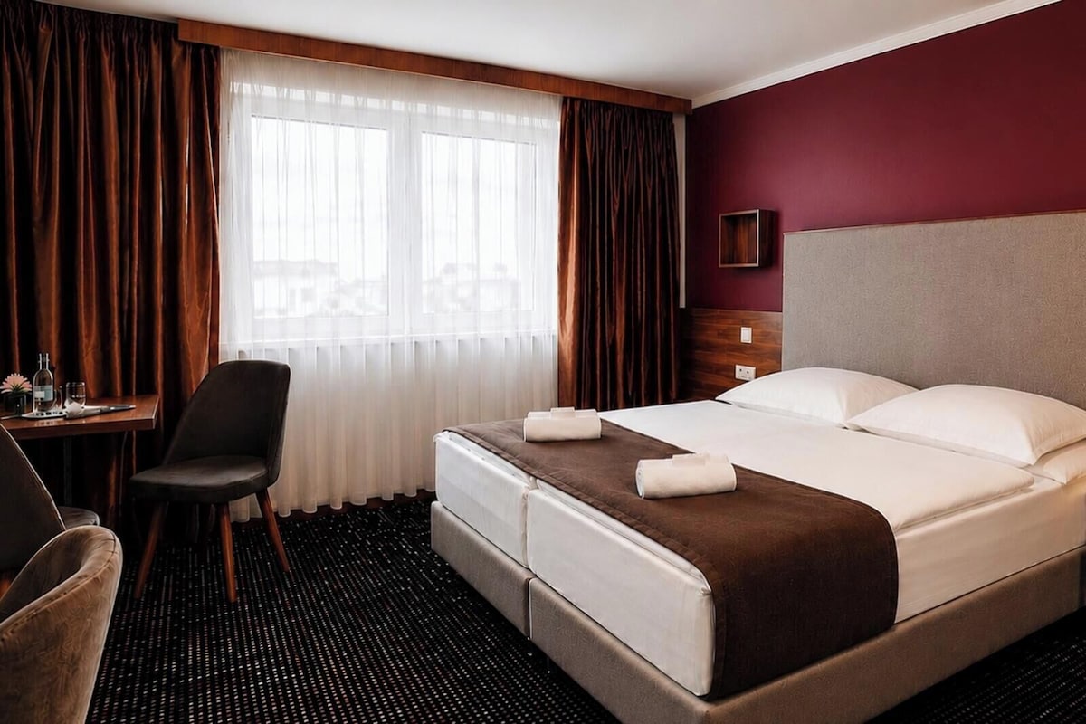 Quarto de hotel com cama, escrivaninha, cadeira e mesa de cabeceira com abajur.