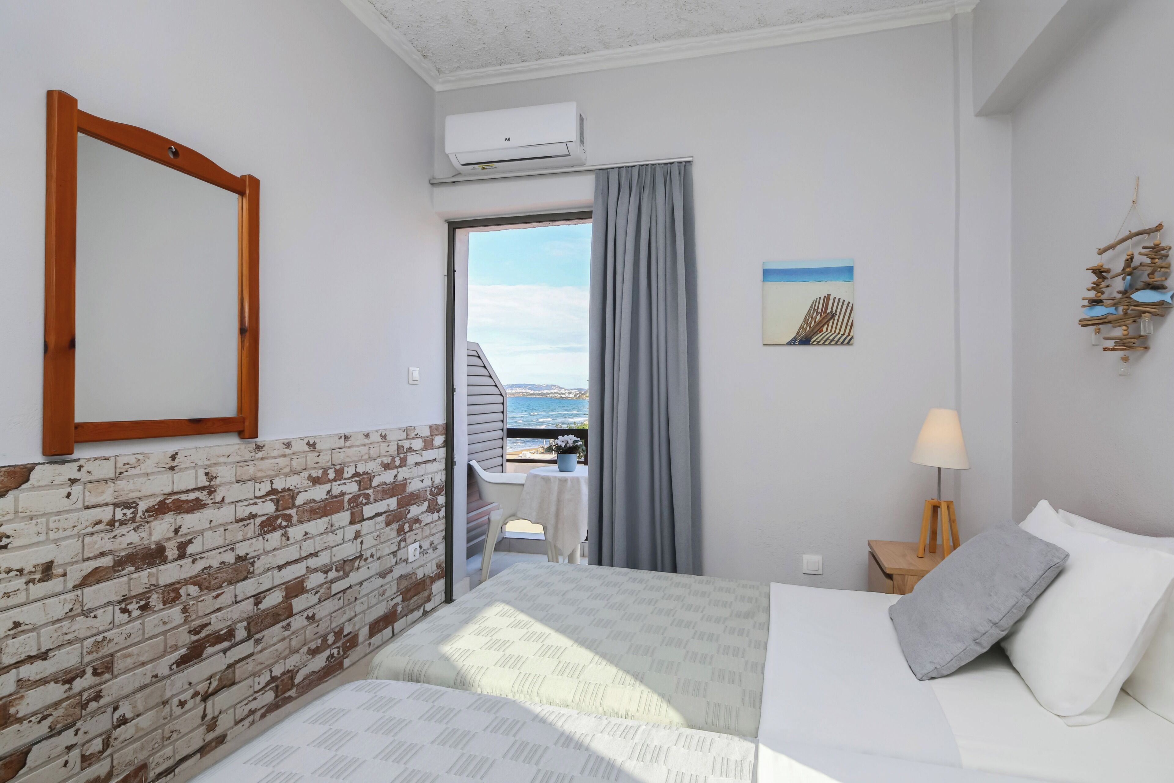 Quarto de hotel com parede de tijolos, cama com roupa de cama branca, espelho com moldura de madeira, janela com vista e uma pequena mesa com abajur.
