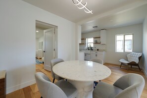 Uma sala de jantar moderna com mesa de mármore e cadeiras cinzentas, cozinha ao fundo e uma grande janela que ilumina naturalmente o espaço.