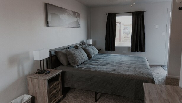 Quarto com cama, mesa de cabeceira, luminária, ventilador de teto, janela com cortinas e uma obra de arte na parede.