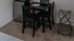 Um quarto pequeno com televisão na parede, micro-ondas, mini-geladeira, mesa de jantar com cadeiras e uma poltrona com encosto.