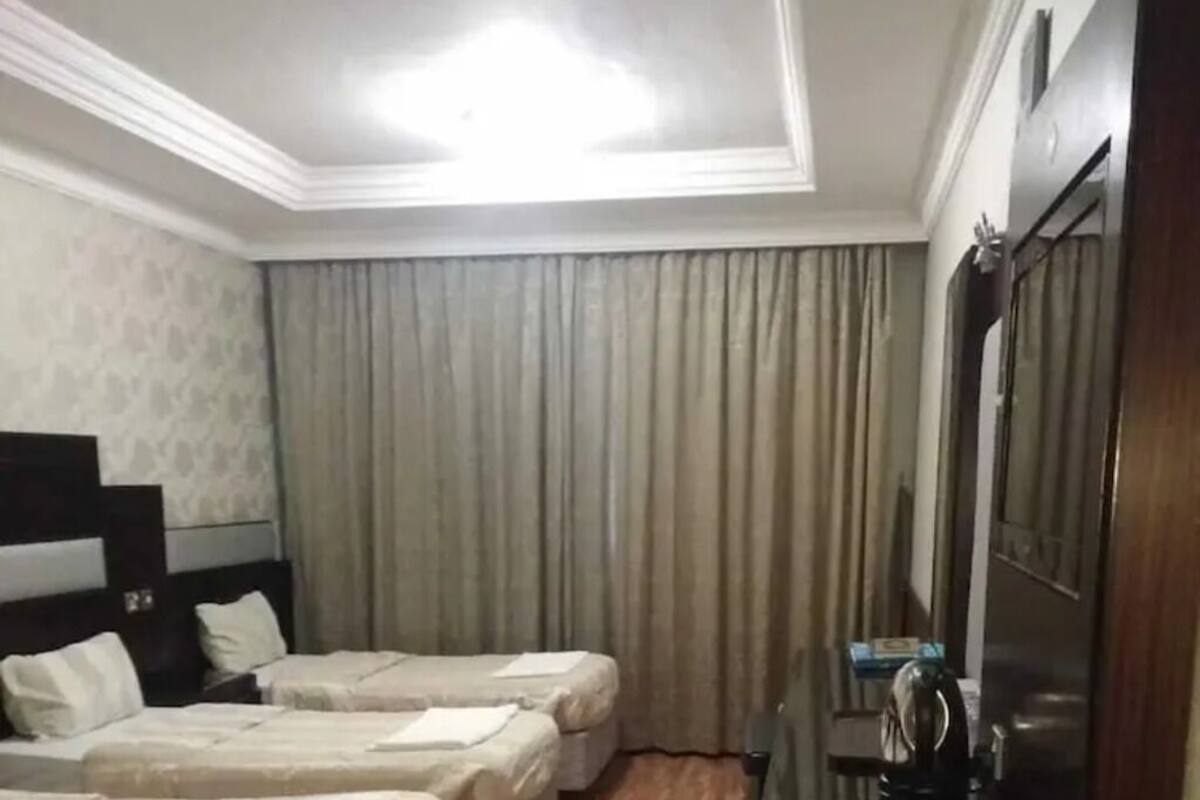 Quarto de hotel com três camas, piso de madeira, uma secretária pequena com espelho e uma lixeira.