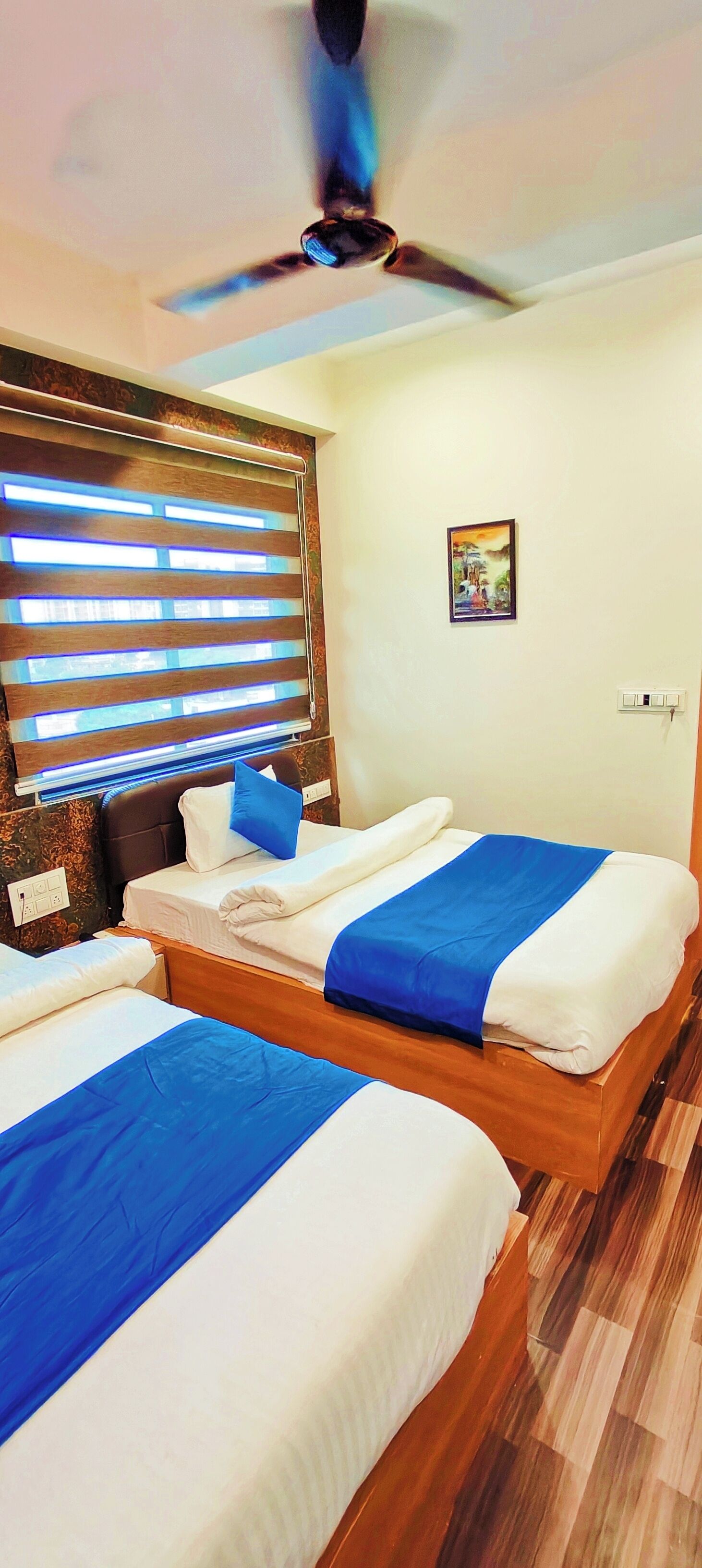 Quarto de hotel com duas camas, ventoinha no teto, janela com persianas, quadro na parede e chão de madeira.