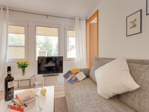 Quarto mobiliado com sofá, televisão, mesa com livros e uma garrafa, e janela com cortinas.