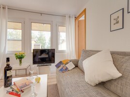 Quarto mobiliado com sofá, televisão, mesa com livros e uma garrafa, e janela com cortinas.