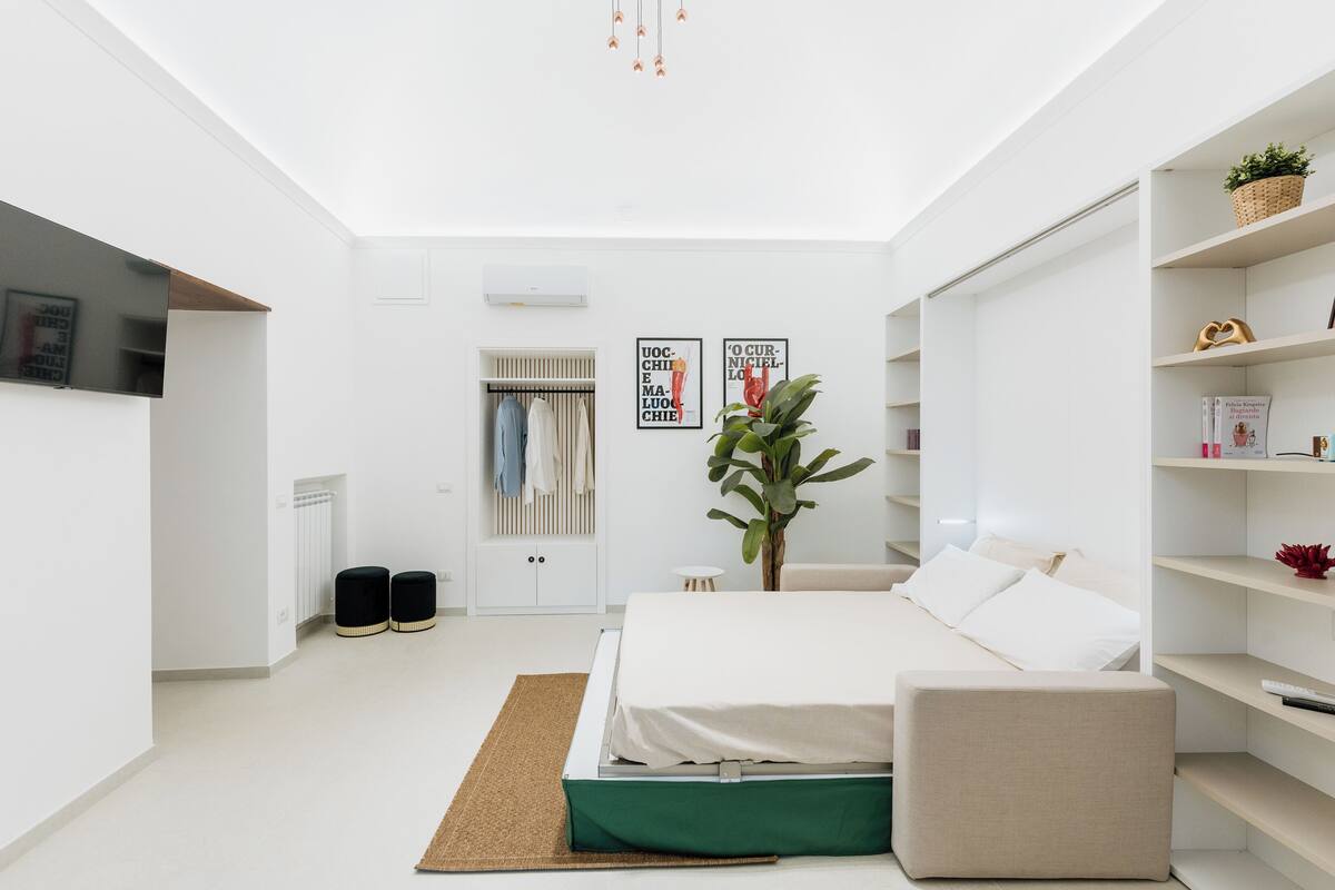 Quarto moderno com cama, um banquinho verde, planta e paredes brancas.