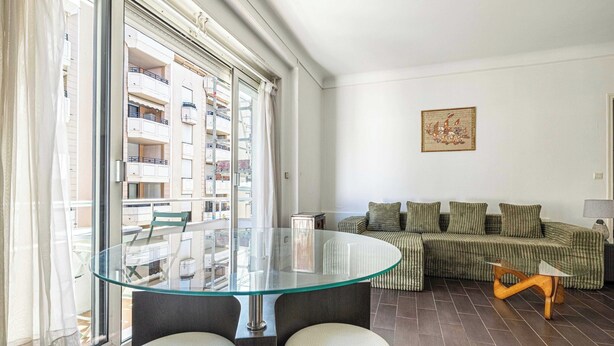 Uma sala de estar moderna com uma mesa de vidro, dois banquinhos estofados, um sofá verde com listras e vista para um edifício residencial através de janelas amplas.