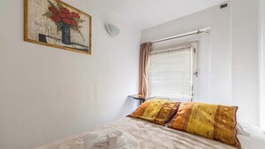 Um quarto com cama, um quadro, janela com cortinas e mesa de cabeceira.