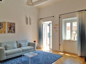Uma sala de estar com sofá, mesa de centro, quadro, porta e janela com cortinas.