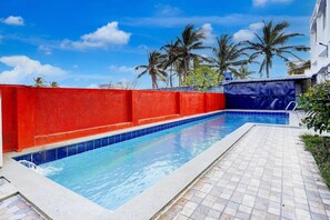 Uma piscina com parede vermelha e azulejos azuis, rodeada por palmeiras e um céu claro.