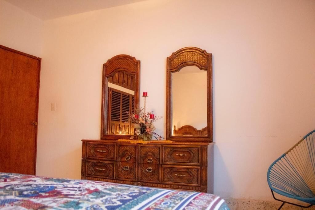 Quarto com cômoda de madeira, cama com roupa de cama estampada, espelho e cadeira azul.