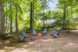 Uma fogueira com quatro cadeiras Adirondack dispostas ao redor em área arborizada.