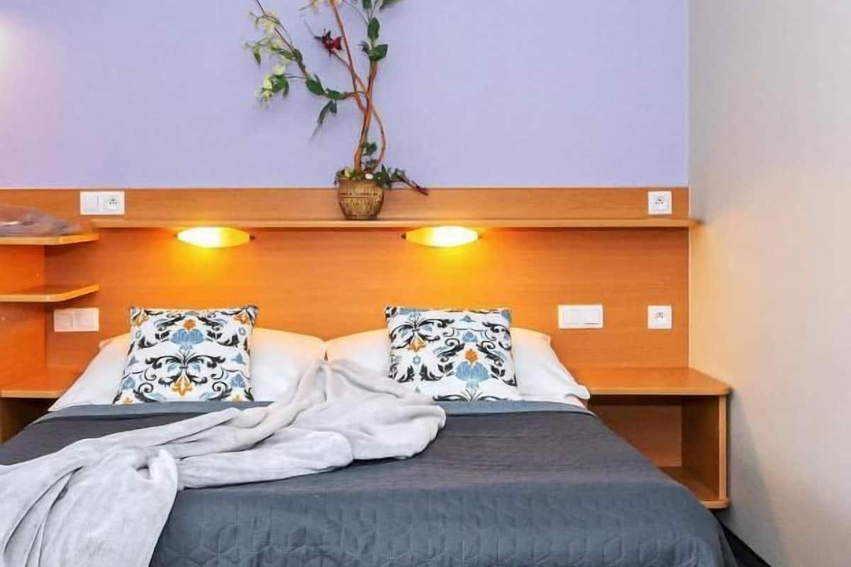 Uma cama de casal com colcha cinza acolchoada, duas almofadas estampadas, um cobertor cinza e uma pequena planta em vaso sobre uma mesinha de cabeceira de madeira.