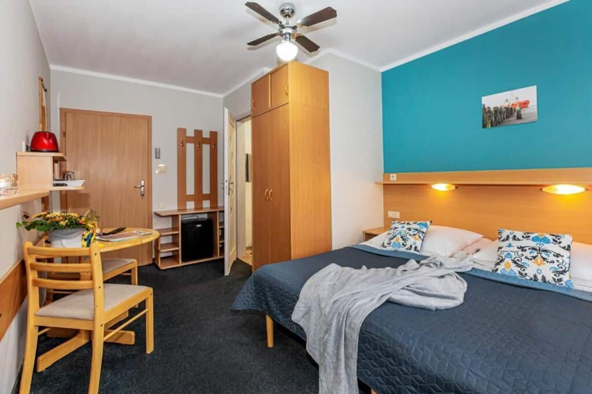Um quarto com cama, escrivaninha, cadeira, guarda-roupa e ventoinha no teto.