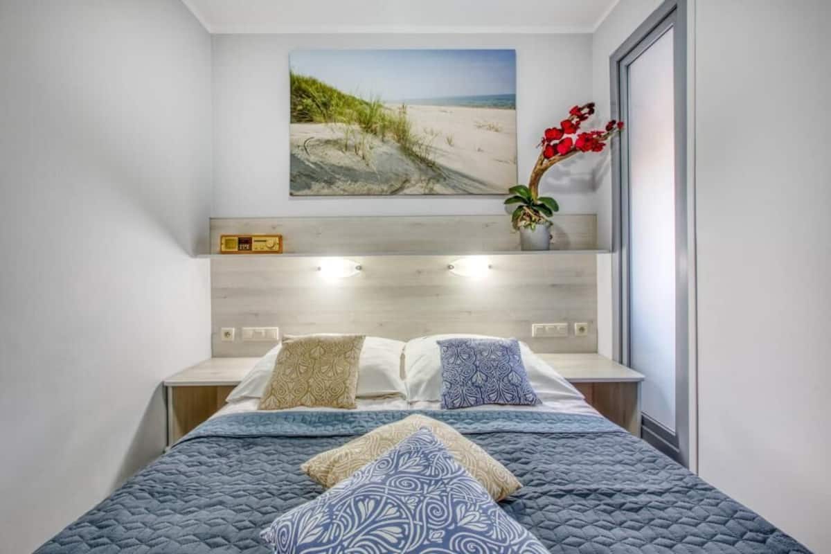 Quarto com cama, mesas de cabeceira, TV fixada na parede, planta e pintura com tema praiano.