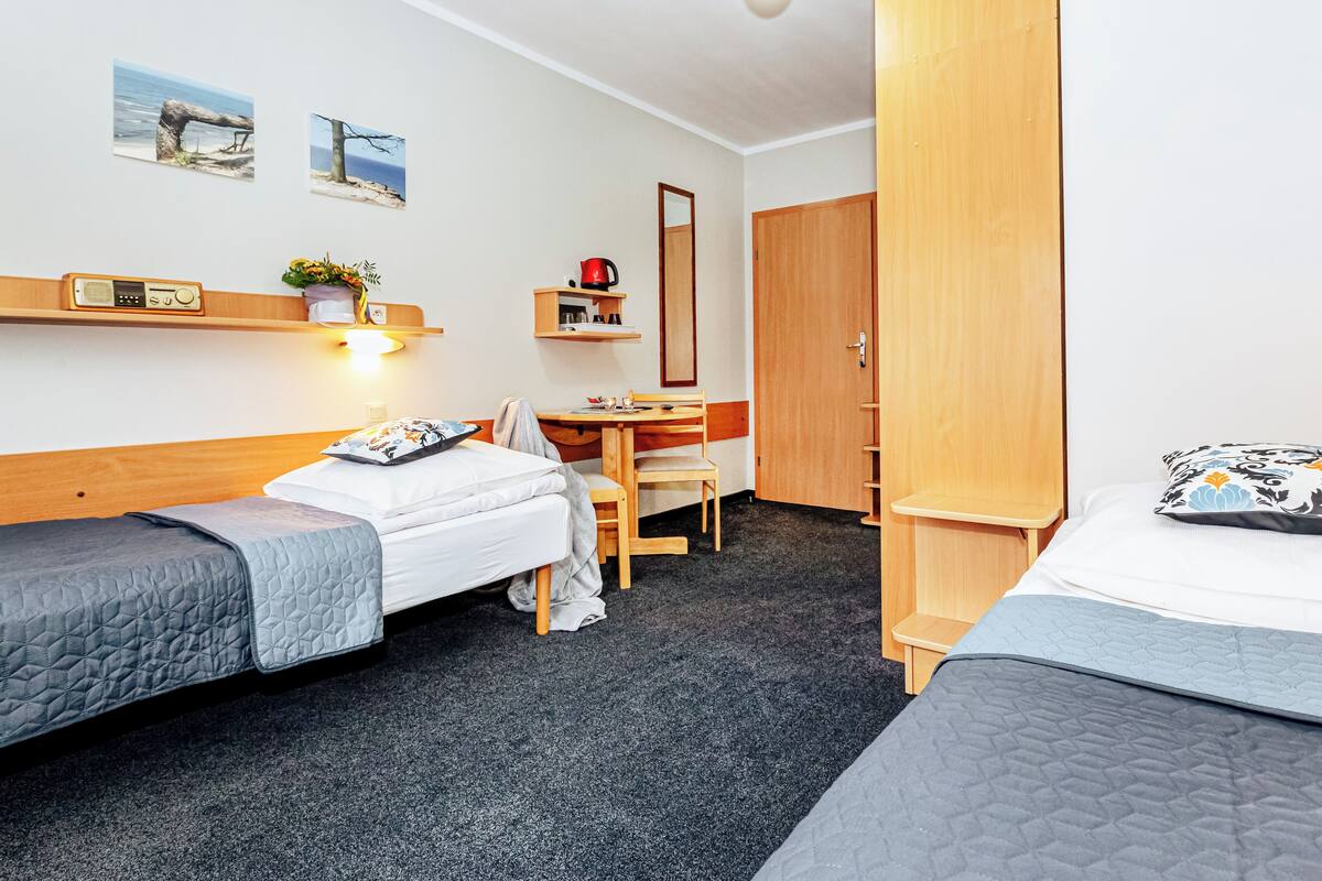 Quarto de hotel com duas camas de solteiro, uma escrivaninha, uma cadeira, um guarda-roupa, ventoinha no teto e obras de arte nas paredes.