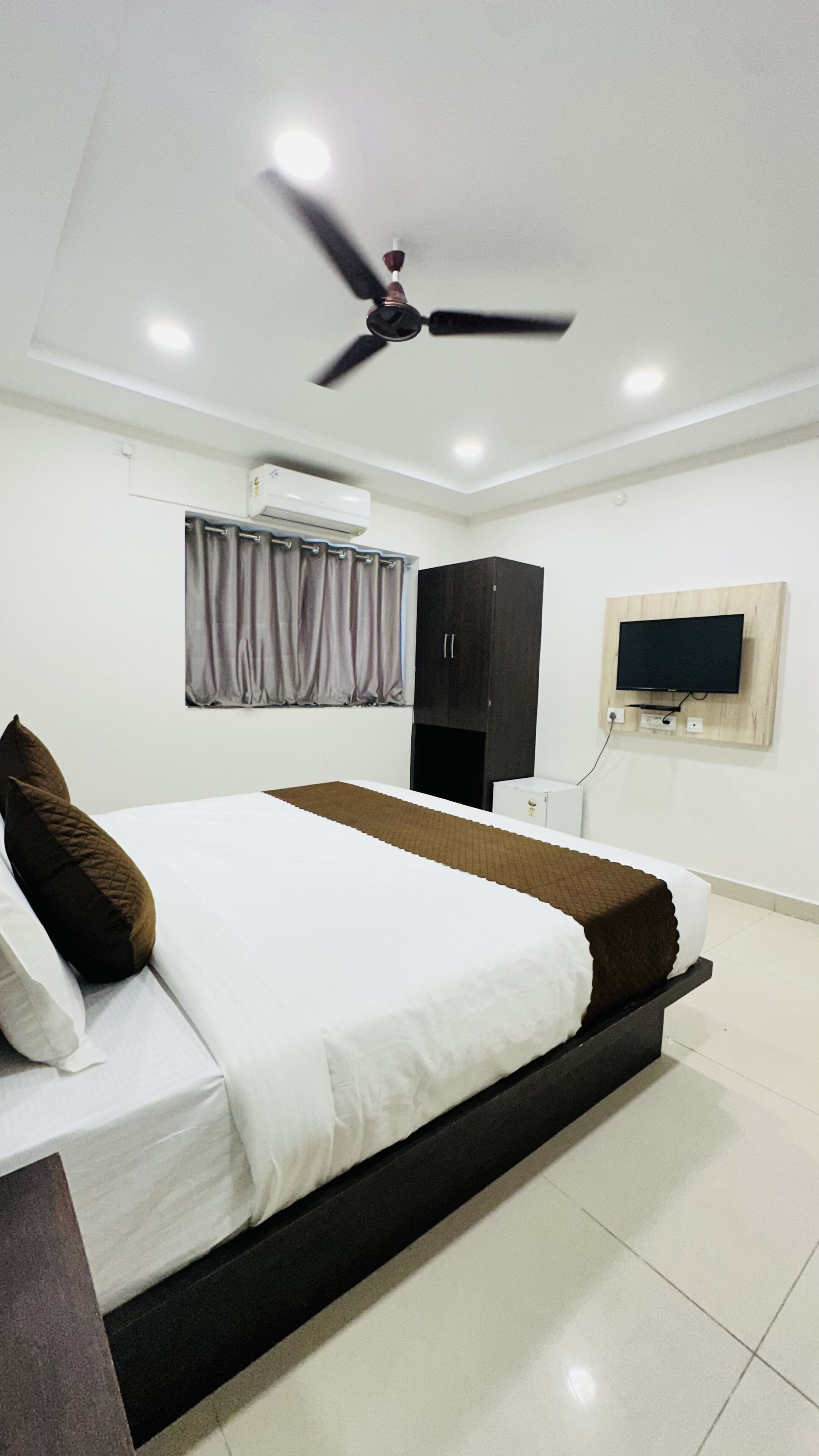 Quarto de hotel moderno com cama grande, ventilador de teto, ar condicionado, guarda-roupa e televisão na parede.