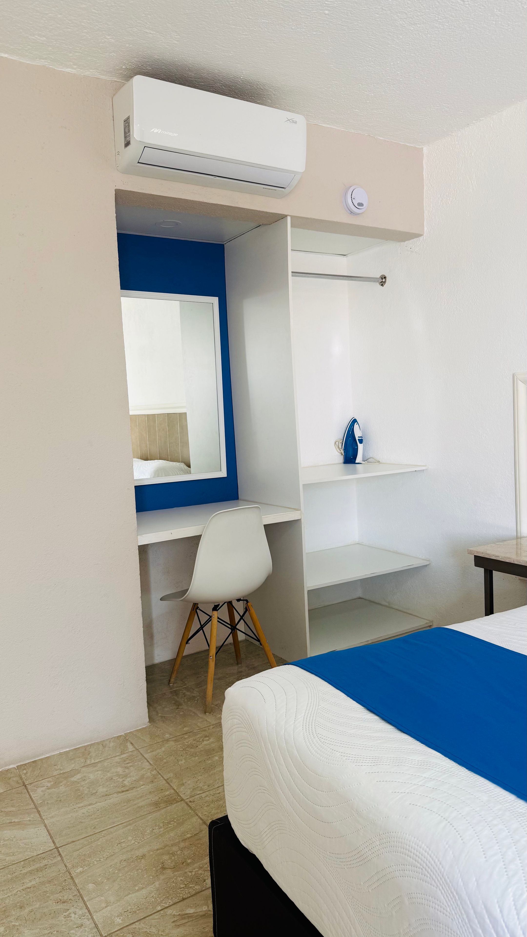 Quarto de hotel com cama, escrivaninha, cadeira, espelho, prateleira e ar condicionado.