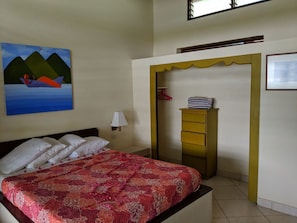 Quarto com cama, quadro, penteadeira e porta que dá para outro cômodo.