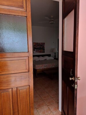 Uma porta de madeira com painel de vidro fosco, um quarto com cama, um ventilador de teto e um quadro na parede.