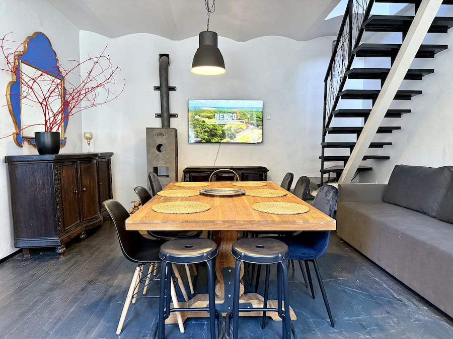 Uma área de refeições com mesa de madeira, cadeiras pretas, luminária pendente preta, uma televisão exibindo 'MEXICO' e uma escada.