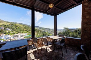 Terraço com mesa e cadeiras, dando vista para um vale com edifícios e vegetação.