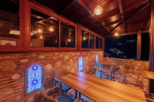 Um restaurante com mesas e cadeiras de madeira, grandes janelas e vista para a cidade à noite.