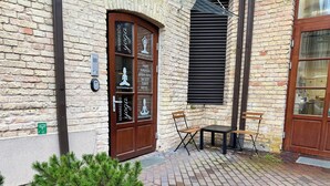 Um edifício de tijolos com uma porta de madeira, uma pequena área externa com mesa e cadeiras, e um vaso de planta.