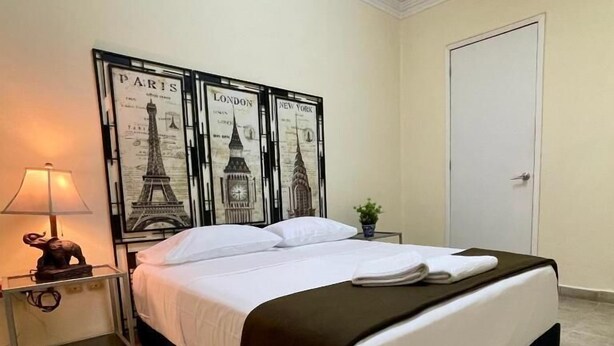 Quarto de hotel com uma cama, mesa de cabeceira com abajur, porta e parede com cartazes de pontos turísticos de Paris.